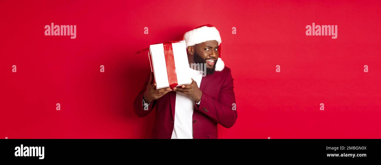 Christmas. Curious Black man in santa hat shaking new year gift, wonder ...