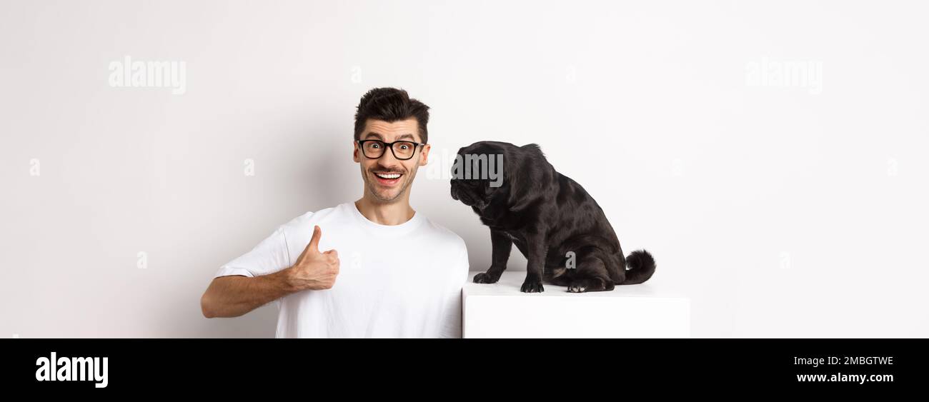 Image of happy and satisfied young man sitting near pug dog and showing ...