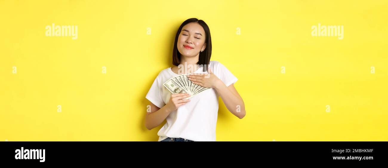 Beautiful asian woman love money, hugging dollars and smiling pleased ...