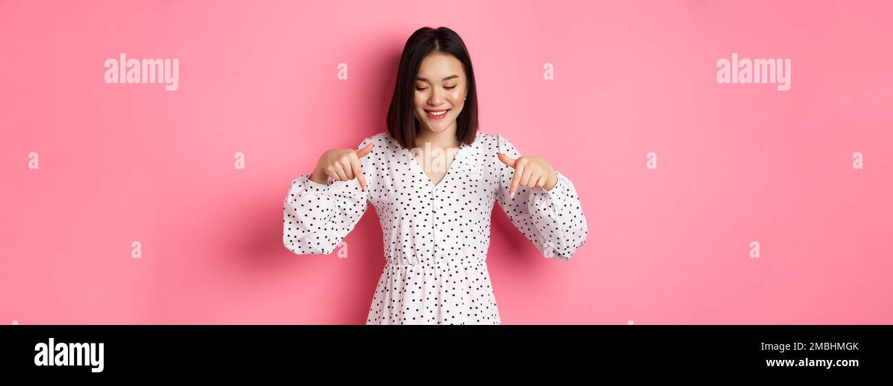 Cute romantic asian woman smiling, pointing fingers and looking down ...