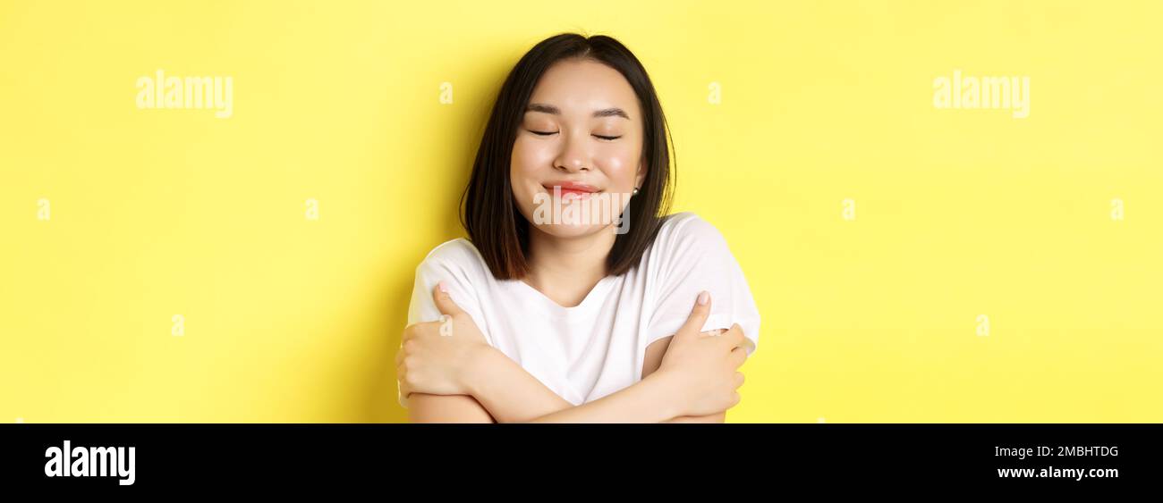 Close up of romantic asian girl hugging herself and dreaming, close ...