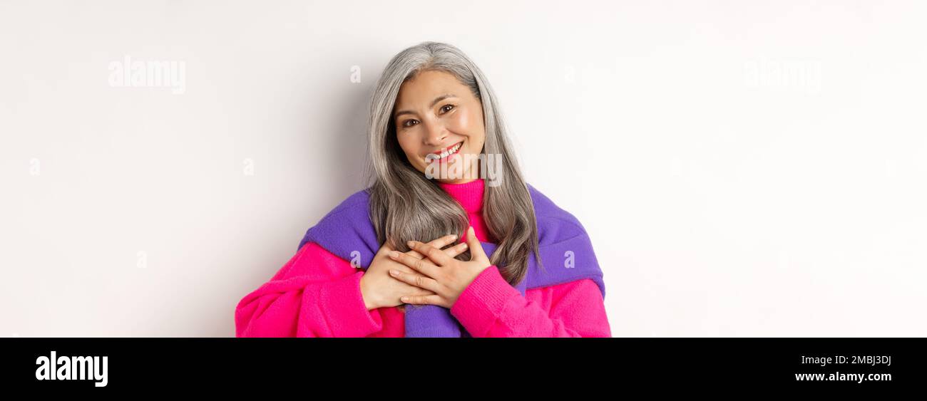 Close up of stylish asian mother holding hands on heart, looking ...