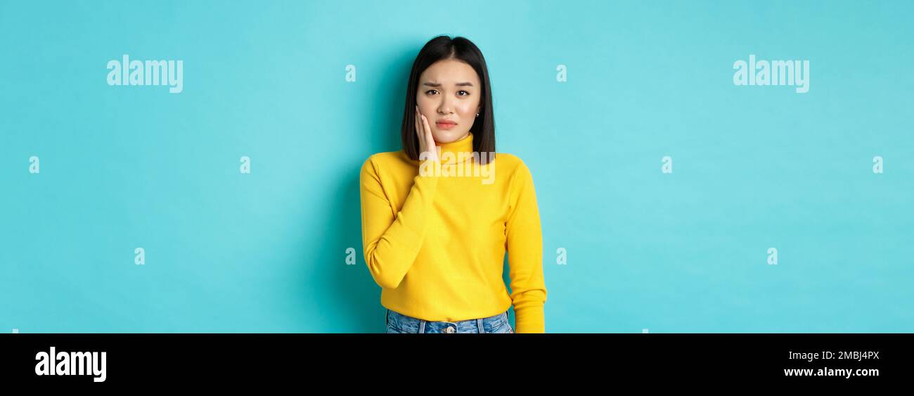 Portrait of asian young woman touching cheek and frowning, looking sad ...