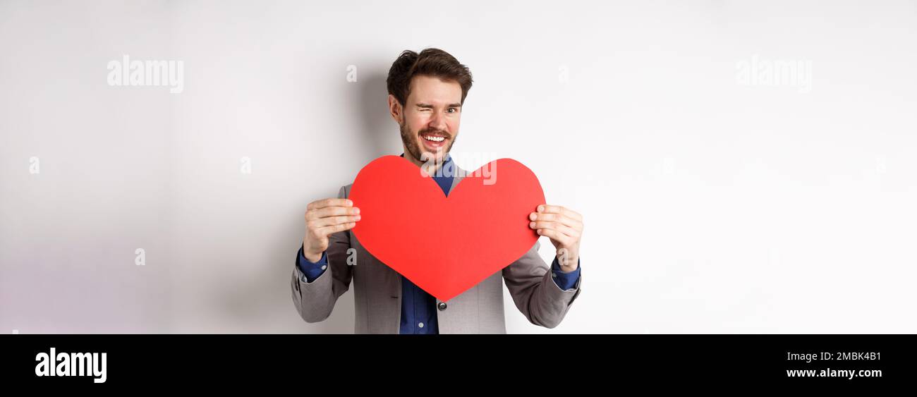 Charismatic young man winking and smiling, showing big red heart cutout ...
