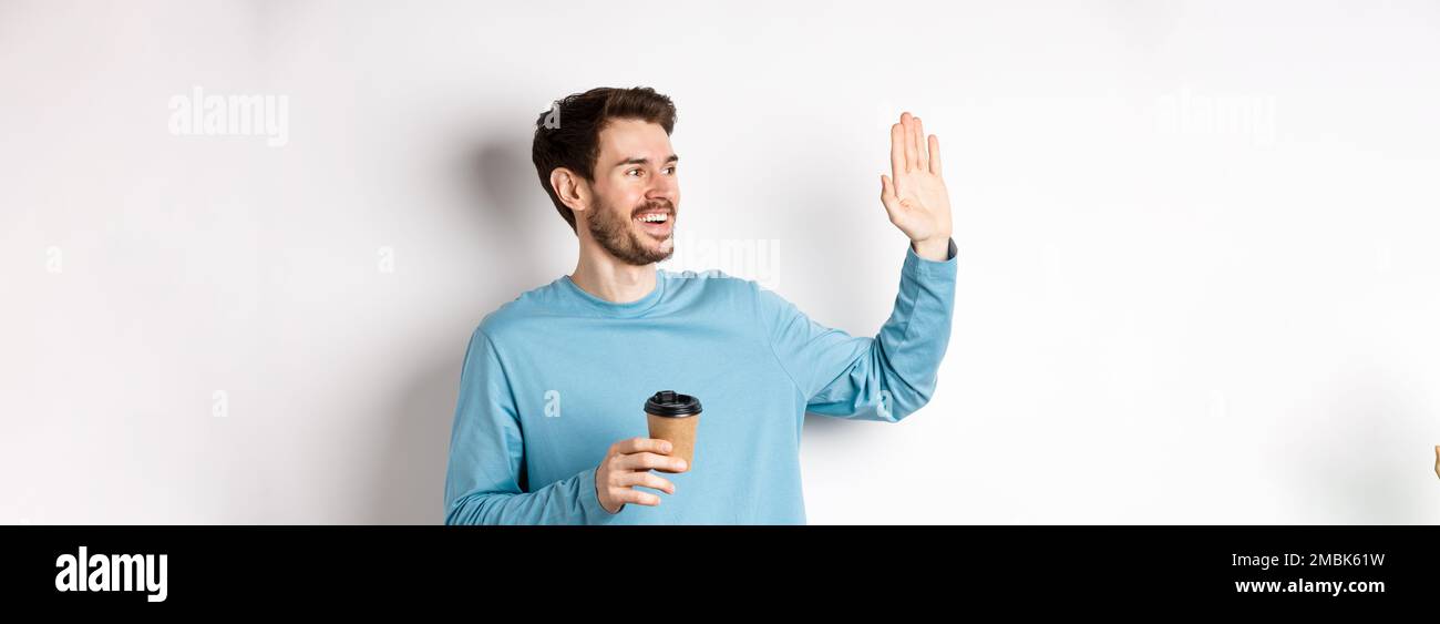 Friendly young man drinking coffee from takeaway cup and saying hello ...