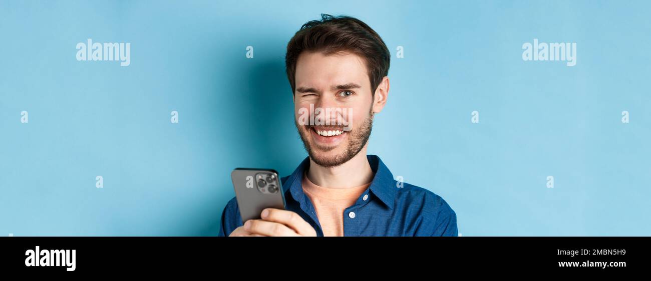 Close up of cheerful man winking and smiling, using mobile phone on ...