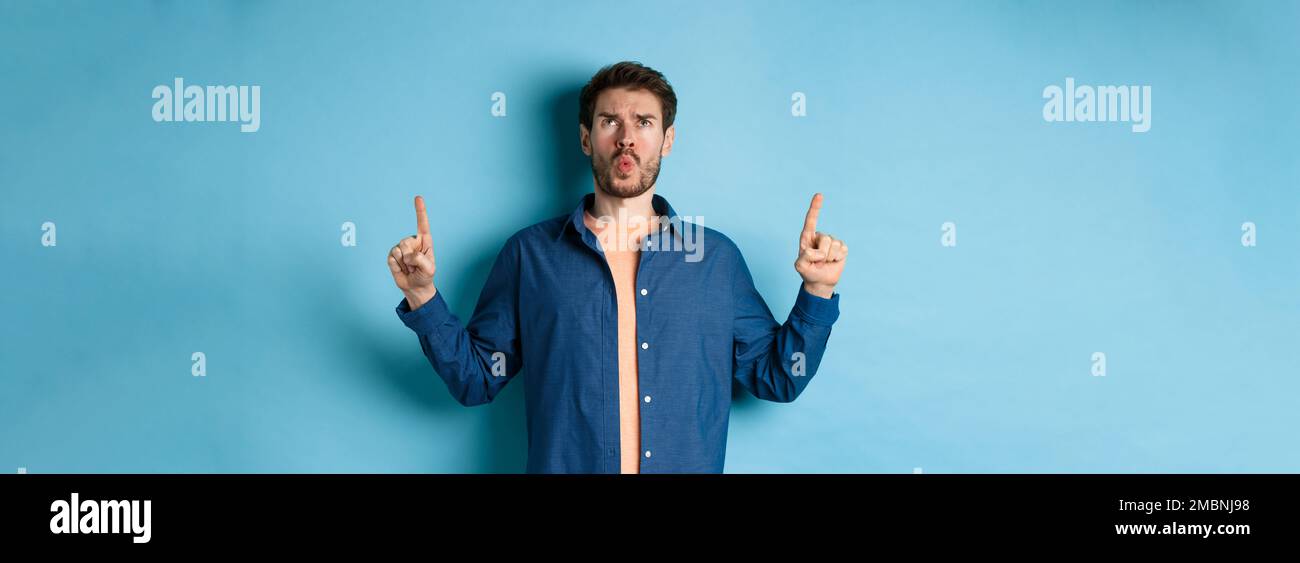 Confused young man frowning, pointing fingers up and looking displeased ...