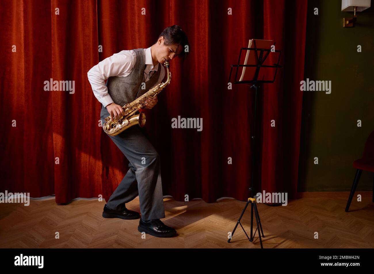 Young attractive musician plays tenor saxophone on stage with red drape ...