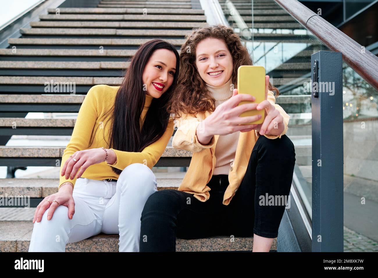 Two Women Smiling While Taking Selfies With A Mobile Phone Sitting On