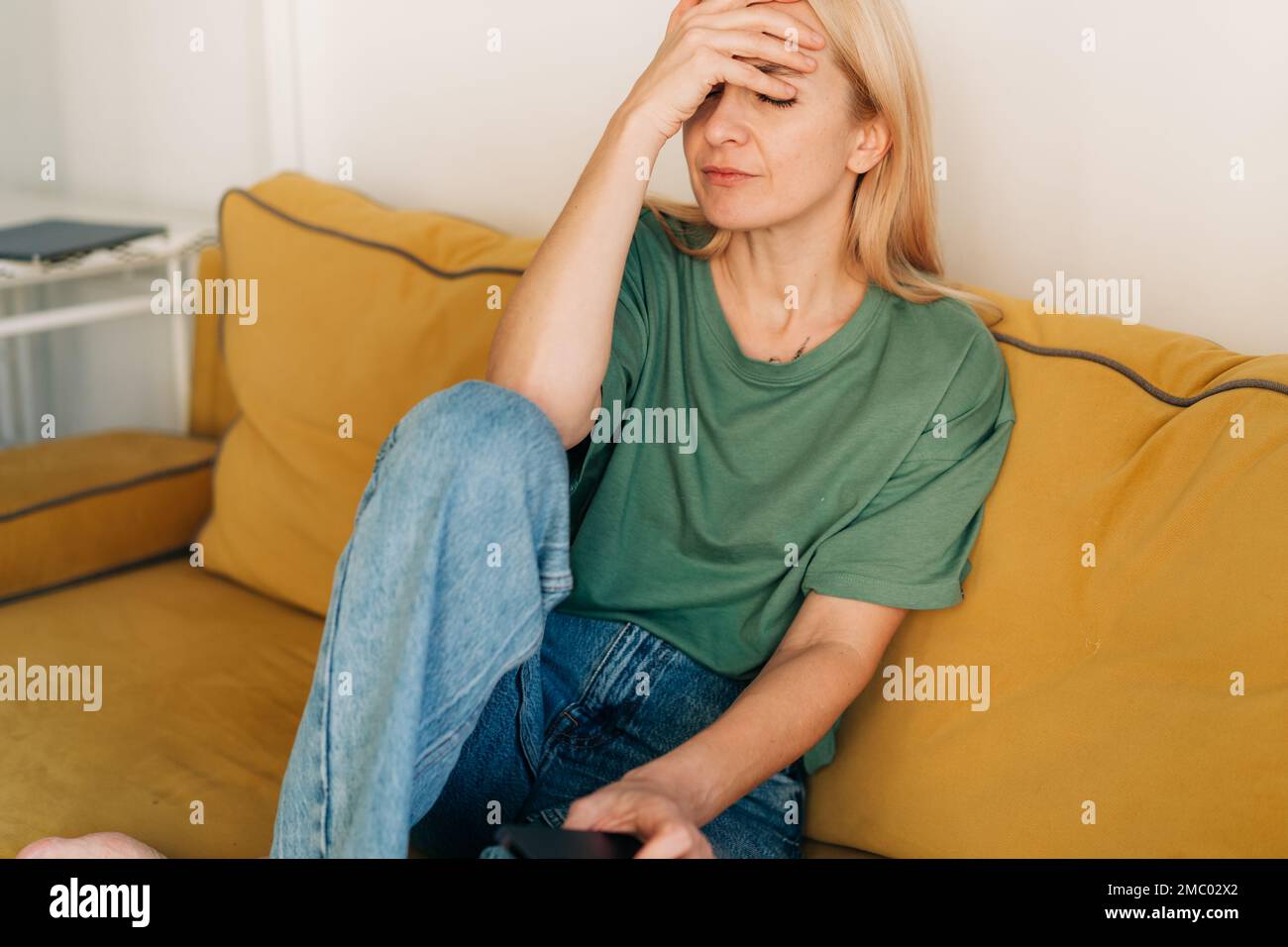 Young sad woman sitting on the couch holds her hand on her sore head ...