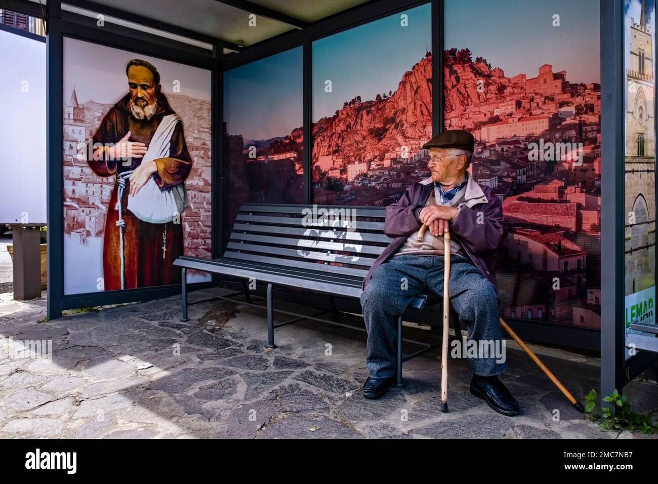 An old man resting on a bench at a bus stop, behind him a poster of the ...