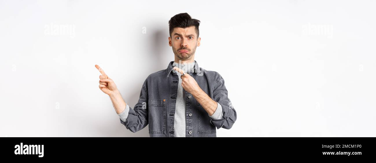 Sad caucasian guy pointing fingers right and pouting, looking hesitant ...