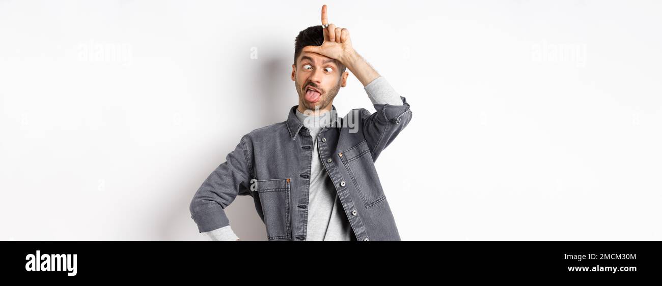 Funny young man making faces and showing tongue with loser sign on ...