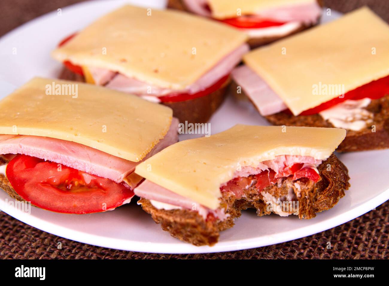 Bitten sandwich with rye bread, tomatoes, ham and cheese on white plate ...