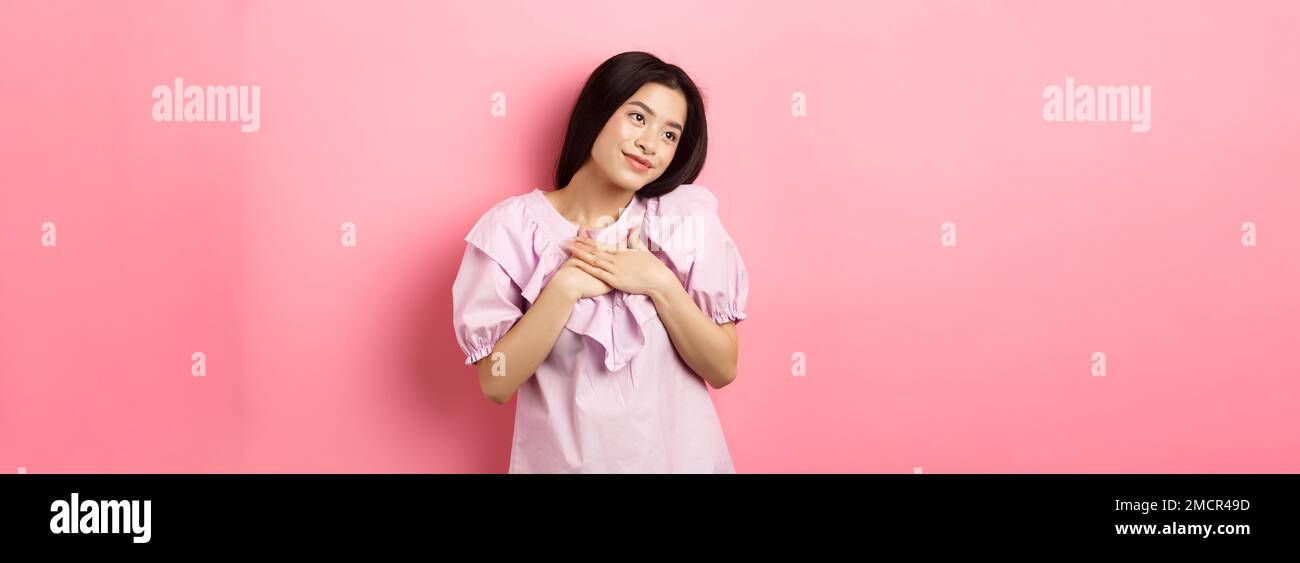 Dreamy asian girl smiling and holding hands on chest, looking left at ...