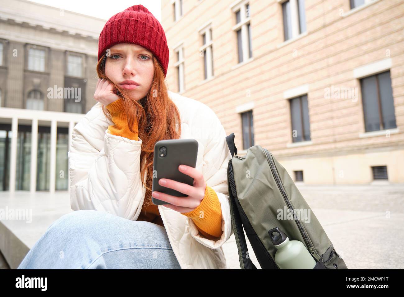 Redhead 20s years girl, sits with smartphone outside building, looks complicated and upset ...
