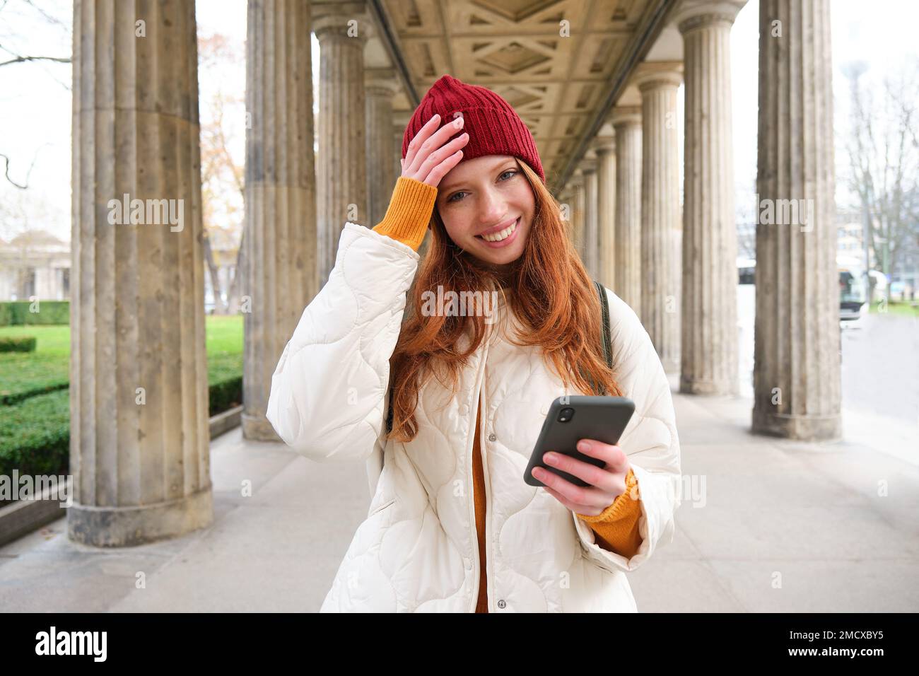 Happy young female model stands on street and holds mobile phone, uses ...
