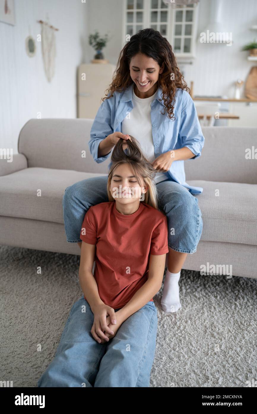 Happy mother and teenage child bonding relaxing together at home, mom braiding daughter's hair ...