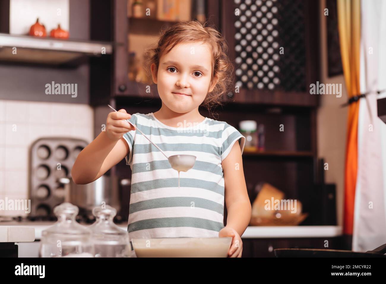Cute little girl kid in the kitchen at home making dough for pancakes ...