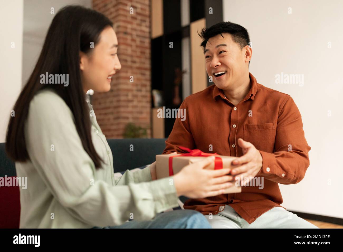 Loving Young Asian Wife Giving Present To Her Happy Mature Husband Sitting On Couch At Home