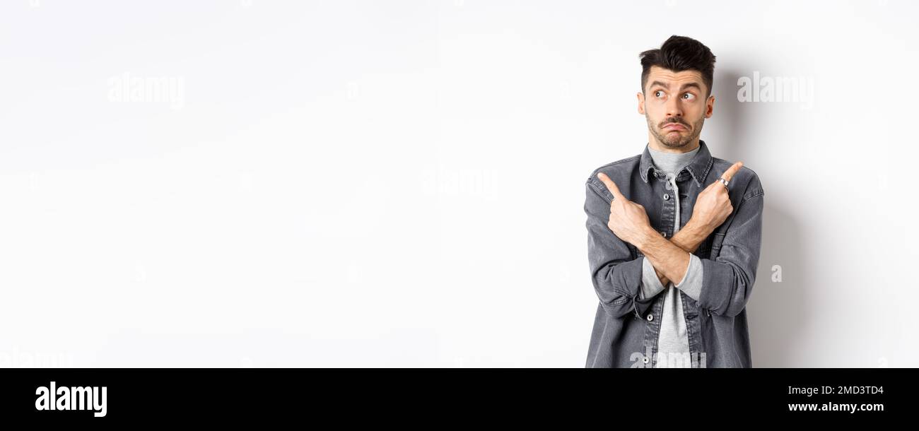 Indecisive handsome guy pointing fingers sideways, looking aside and ...