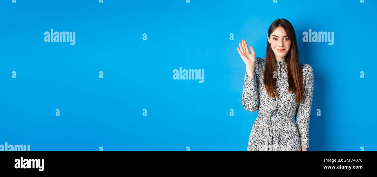 Cute smiling woman waving hand to greet you, saying hello with shy grin ...