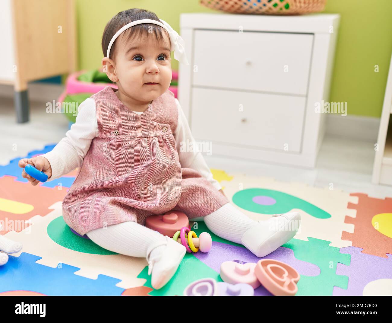 Adorable hispanic baby sitting on floor holding toy at kindergarten ...