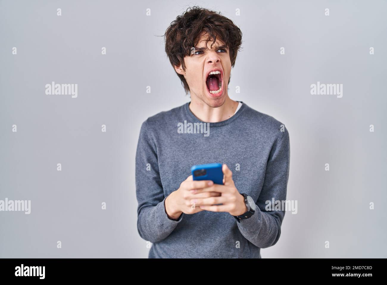 Young man using smartphone typing a message angry and mad screaming ...