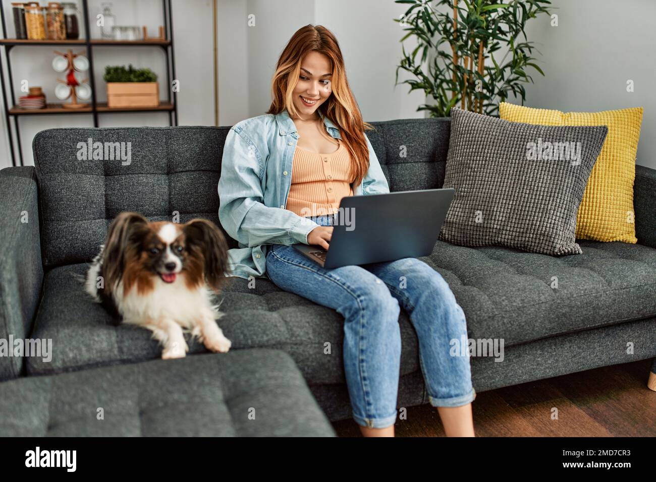 Young caucasian woman using laptop sitting on sofa with dog at home ...