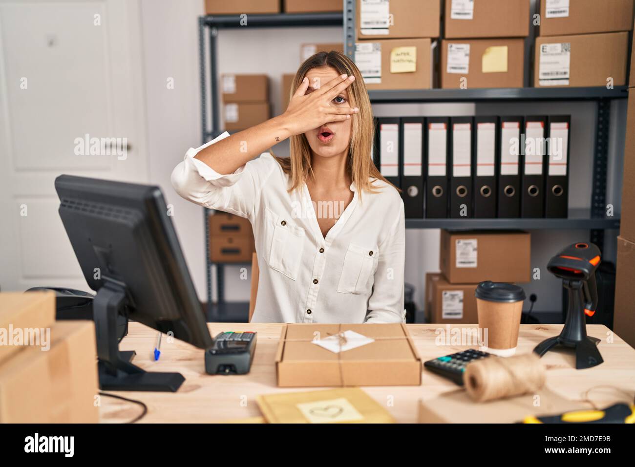 Young blonde woman working at small business ecommerce peeking in shock ...