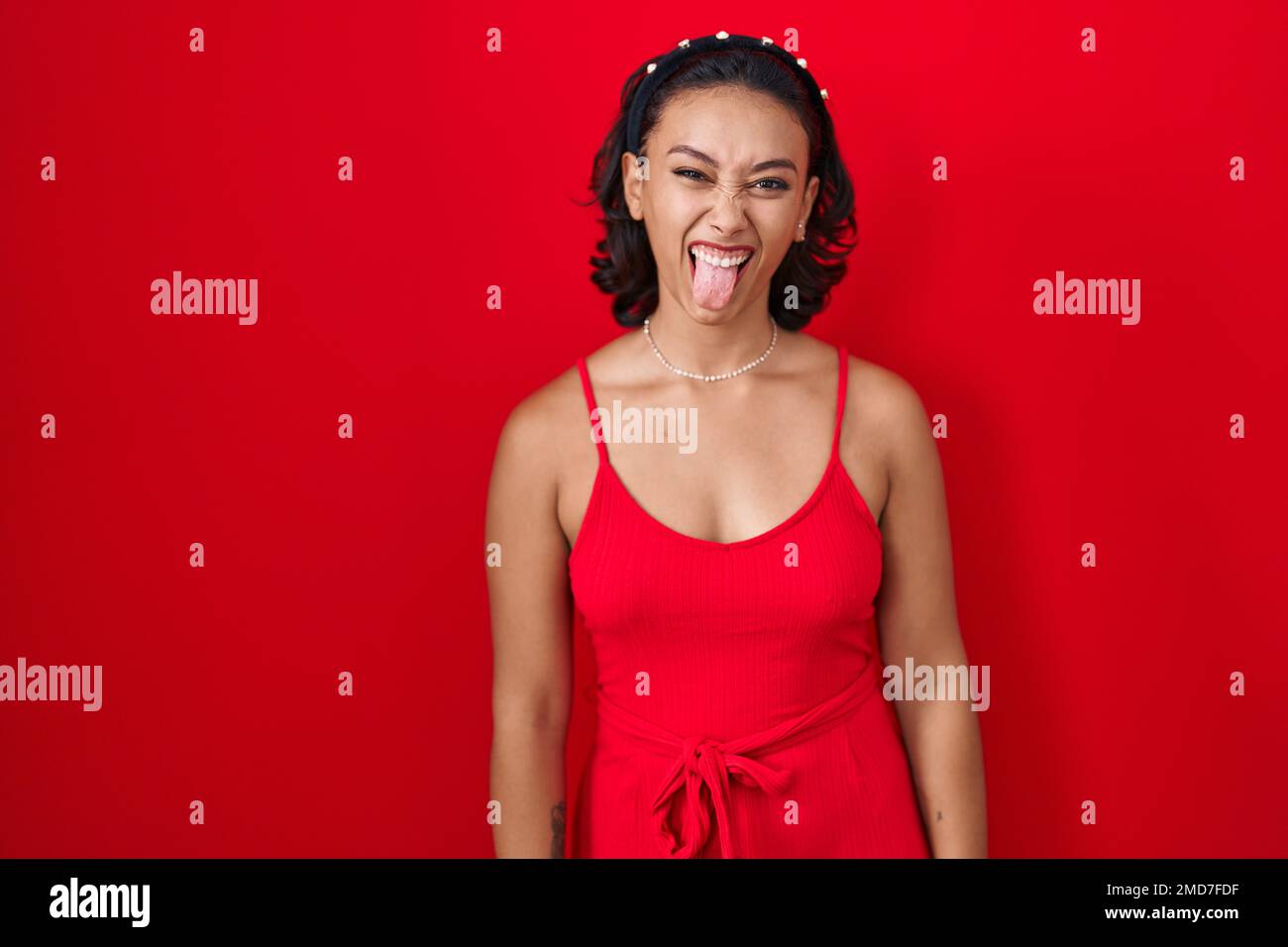 Young hispanic woman standing over red background sticking tongue out ...