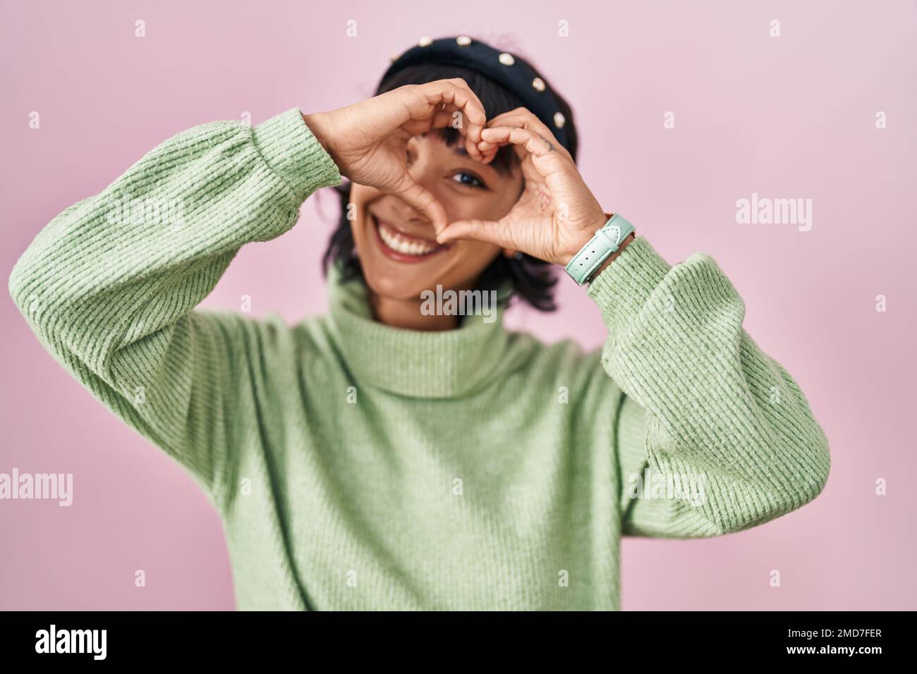 Young beautiful woman standing over pink background doing heart shape ...