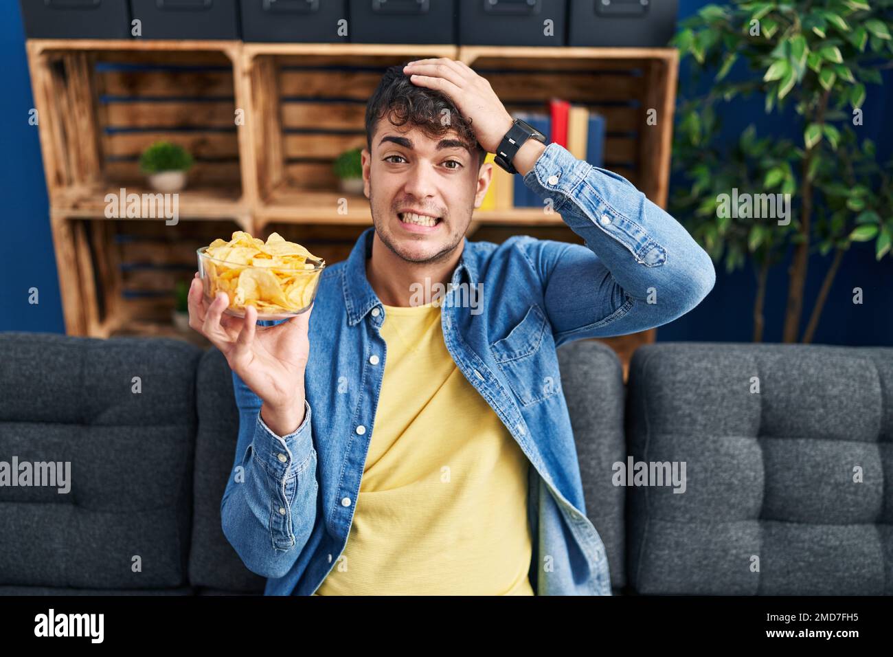 Young hispanic man holding potato chips stressed and frustrated with ...