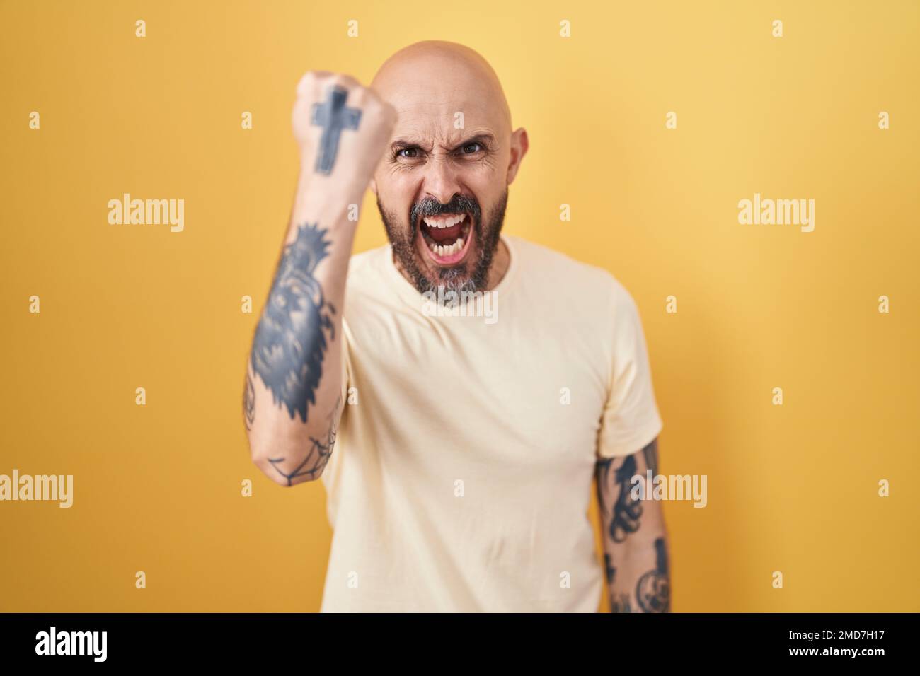 Hispanic man with tattoos standing over yellow background angry and mad ...