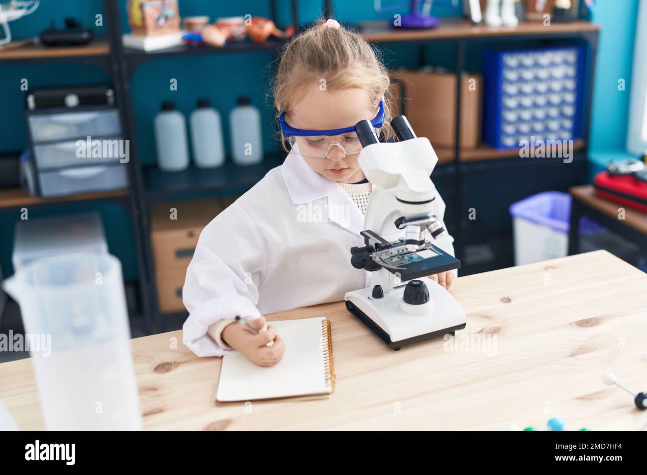 Adorable blonde girl student using microscope writing on notebook at ...