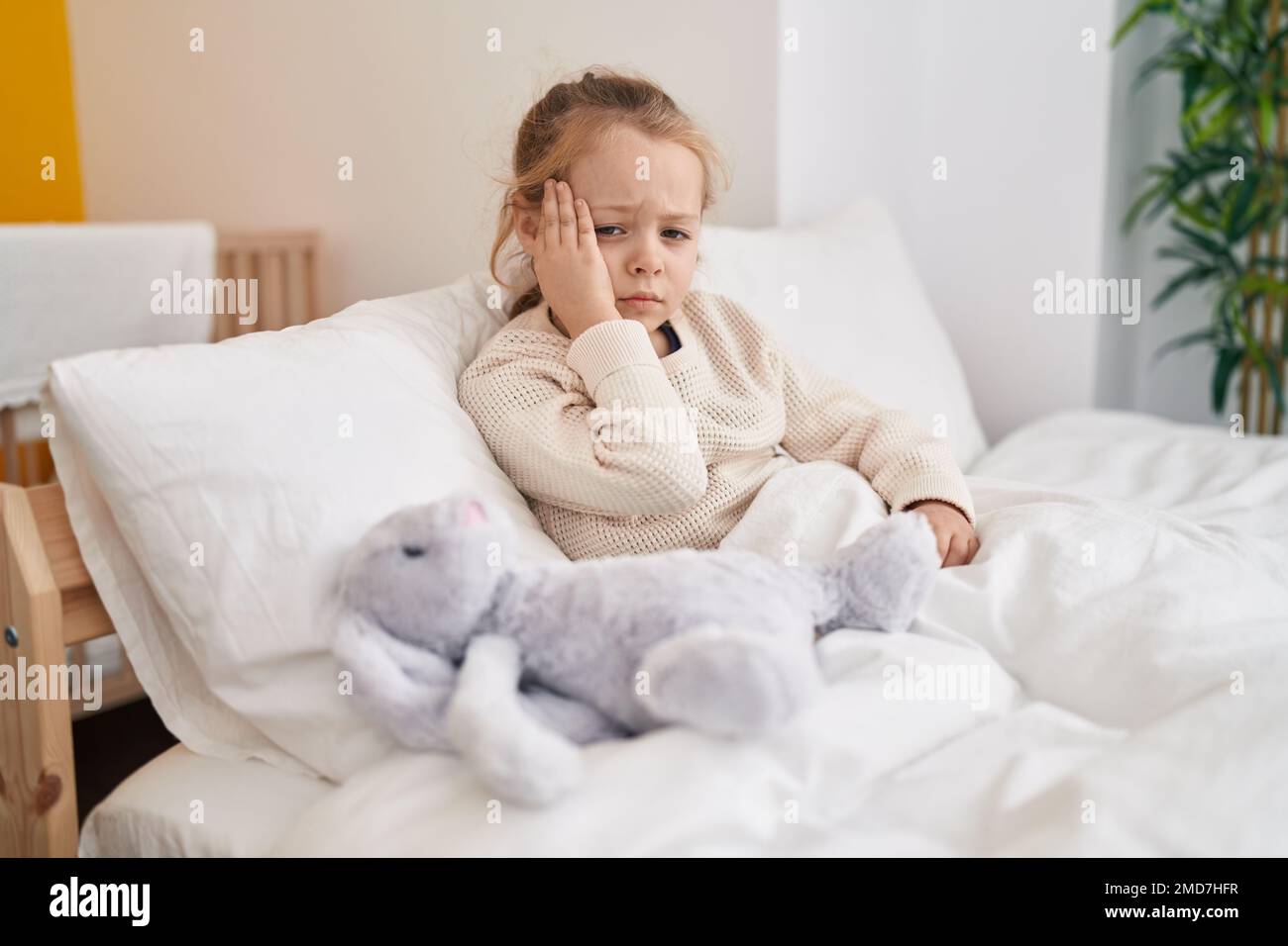 Adorable blonde girl touching head for headache sitting on bed at ...