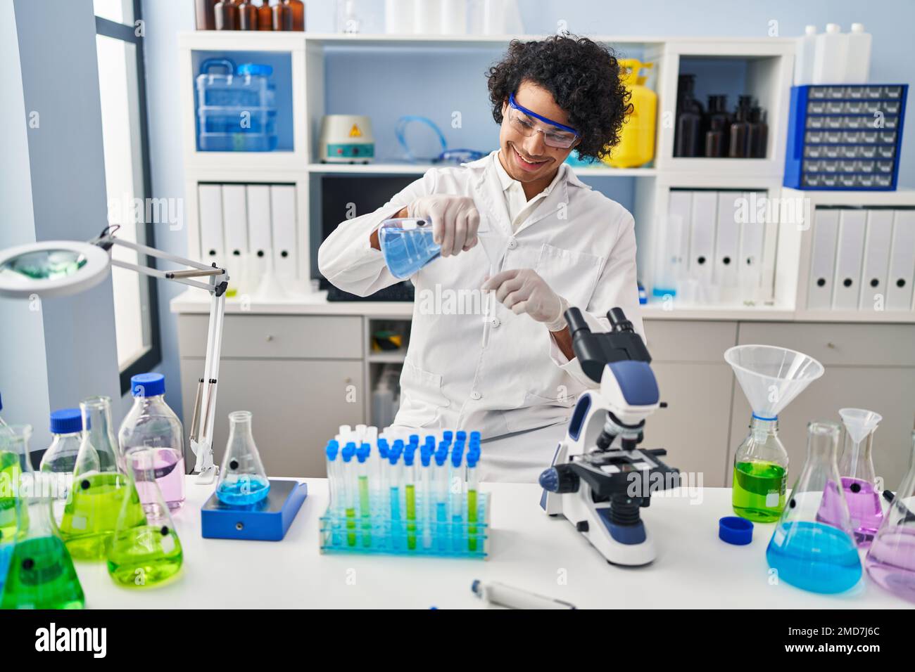 Young hispanic man wearing scientist uniform pouring liquid on test ...