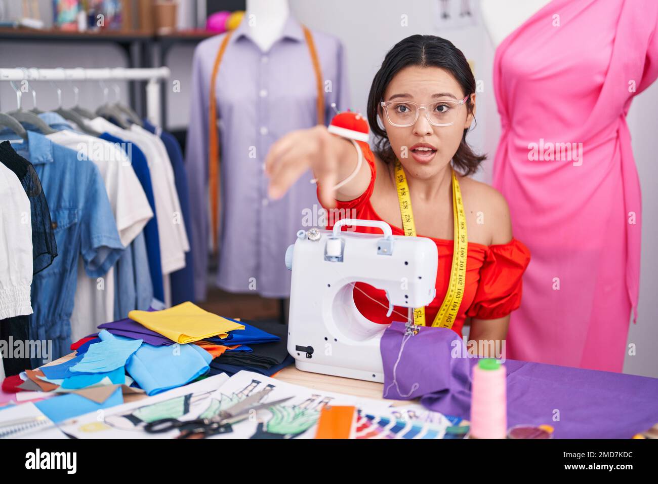 Hispanic young woman dressmaker designer using sewing machine pointing ...