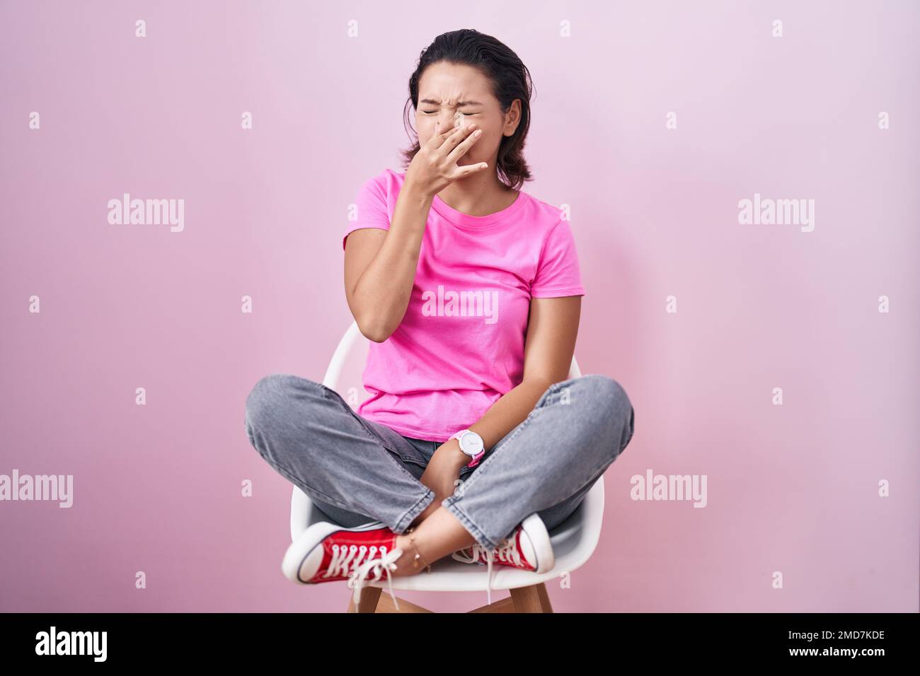 Hispanic young woman sitting on chair over pink background smelling ...