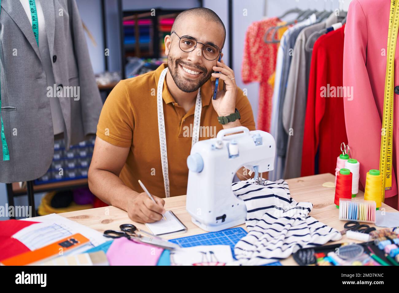Young latin man tailor talking on smartphone writing on notebook at ...