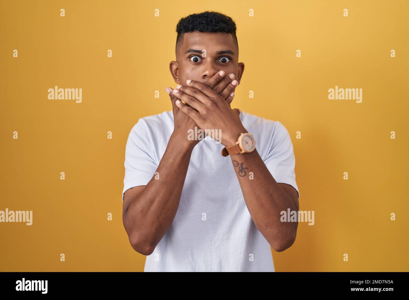 Young hispanic man standing over yellow background shocked covering ...