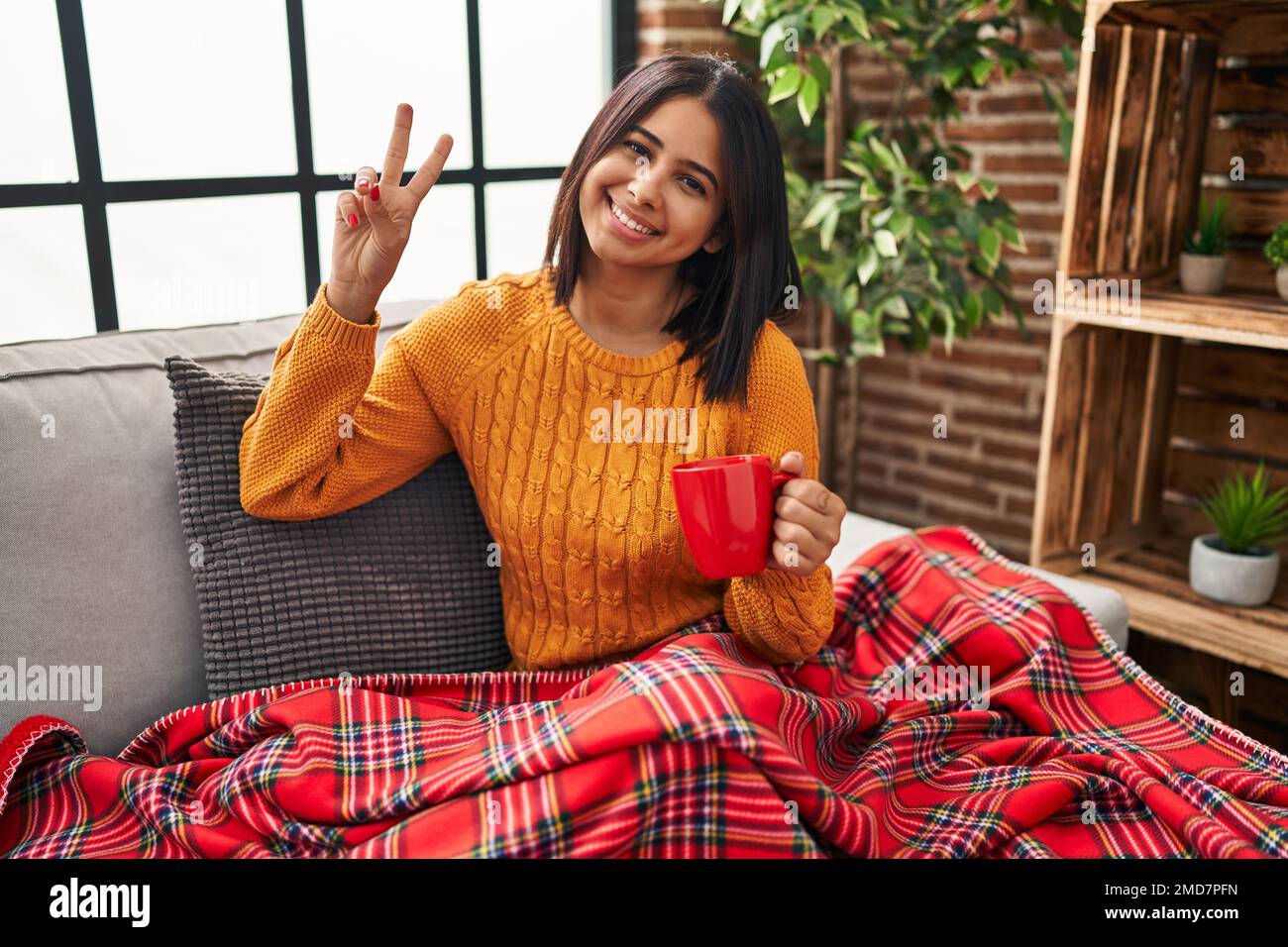 Young hispanic woman sitting on the sofa drinking a coffee at home ...