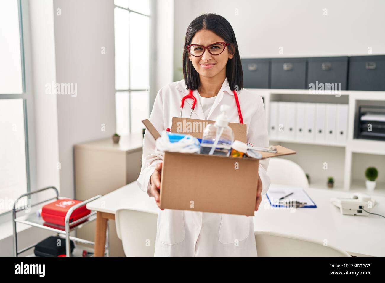 Young hispanic doctor woman holding box with medical items clueless and ...