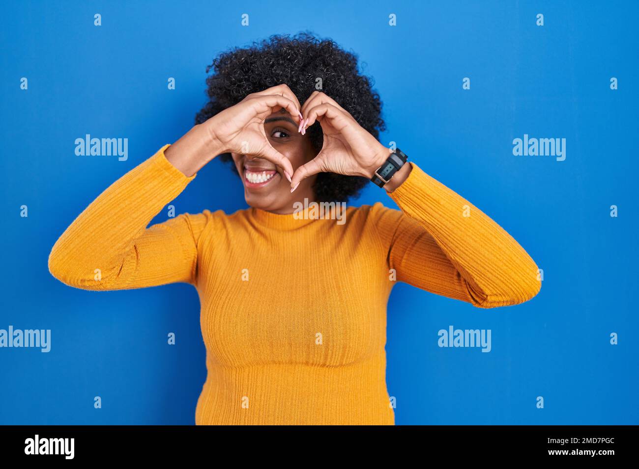 Black woman with curly hair standing over blue background doing heart ...