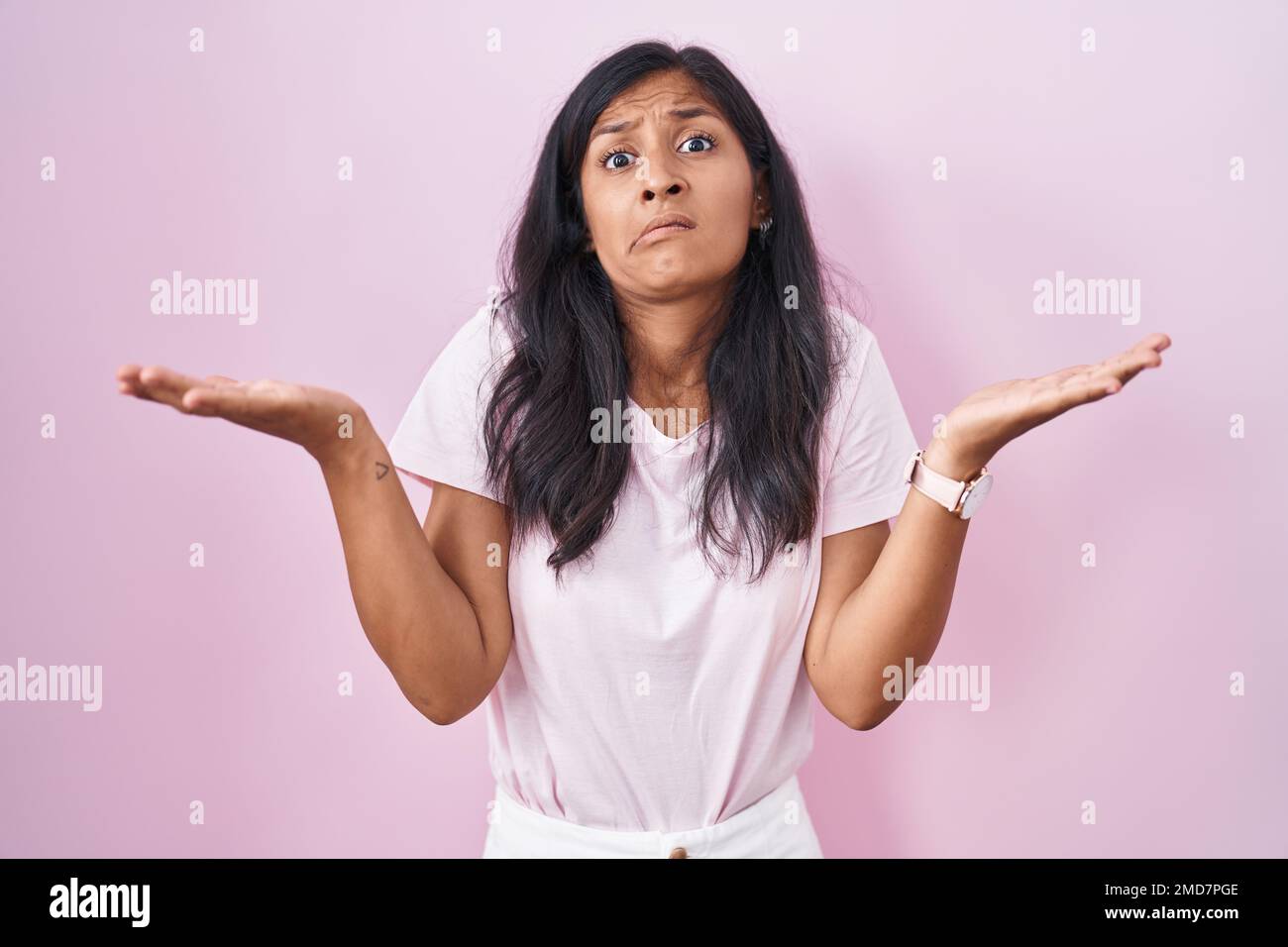 Young hispanic woman standing over pink background clueless and ...