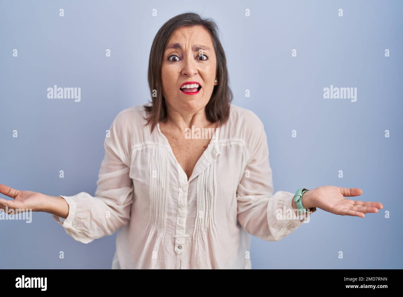 Middle age hispanic woman standing over blue background clueless and ...
