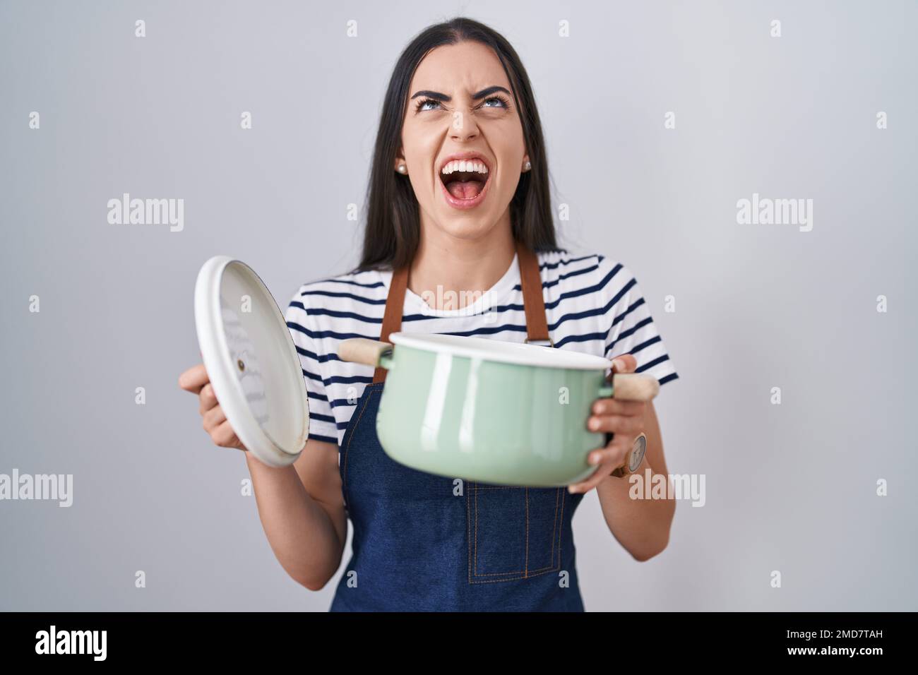 Young brunette woman wearing apron holding cooking pot angry and mad ...