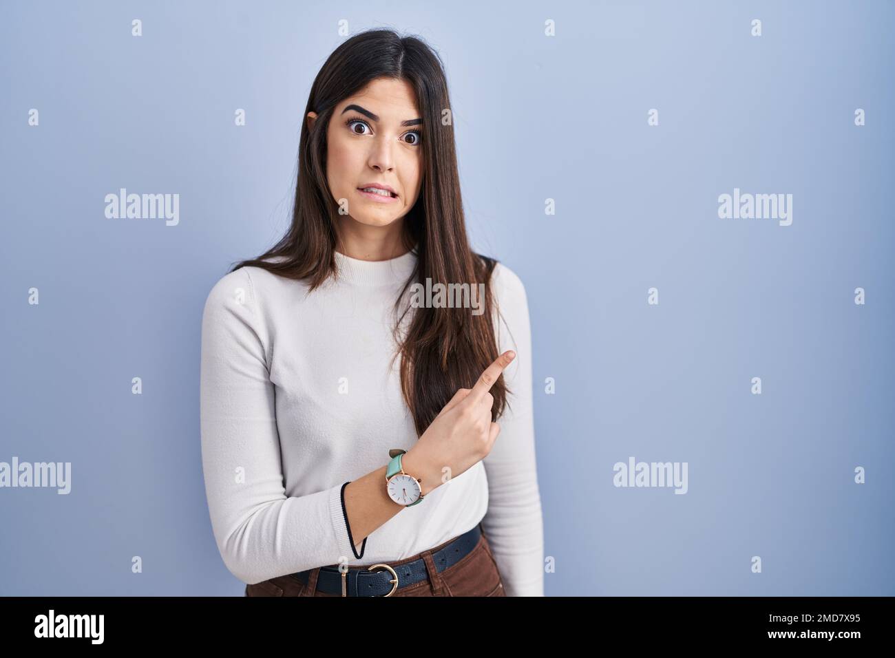 Young brunette woman standing over blue background pointing aside ...