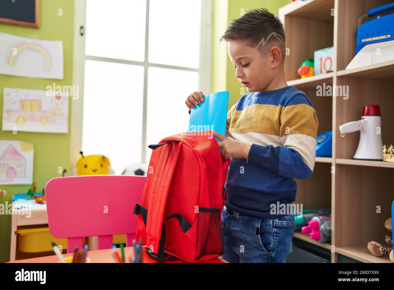 Adorable hispanic boy student holding book of backpack at kindergarten ...