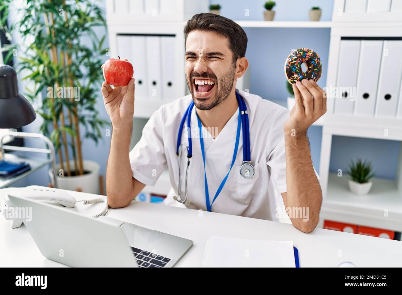 Young hispanic dietitian man holding doughnut and apple angry and mad ...